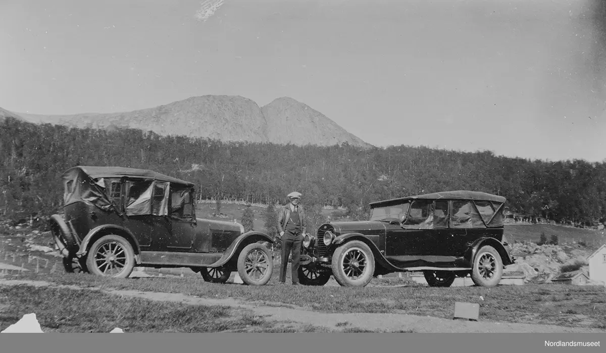 2 biler. Mellom bilene står en mann, kledd i mørke klær og lue ...