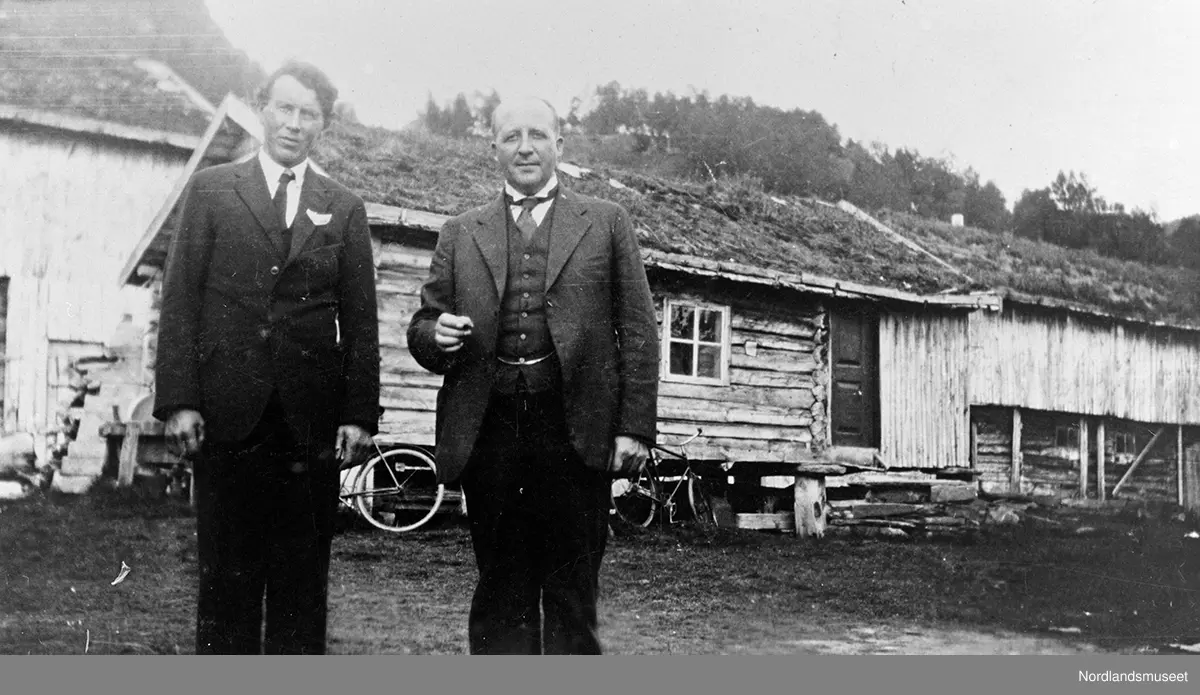 To menn kledd i penklær, stabbur og fjøs i bakgrunnen. Johan Brekke og ...