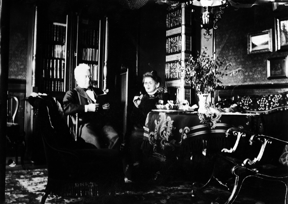 Abraham G. og Claudia Schweigaard i stuen sin, Han leser og hun spiser ...