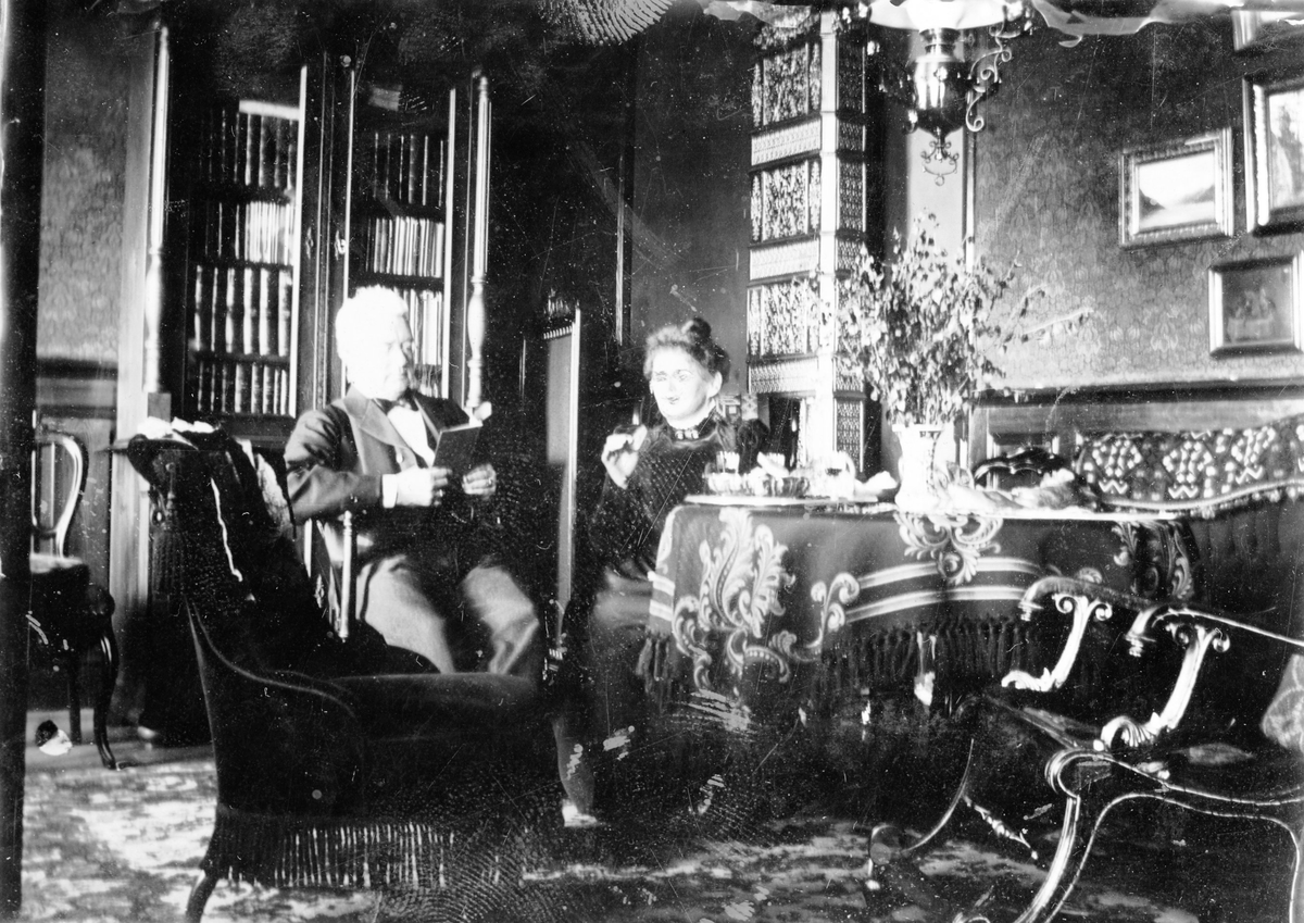 Abraham G. og Claudia Schweigaard i stuen sin, Han leser og hun spiser ...