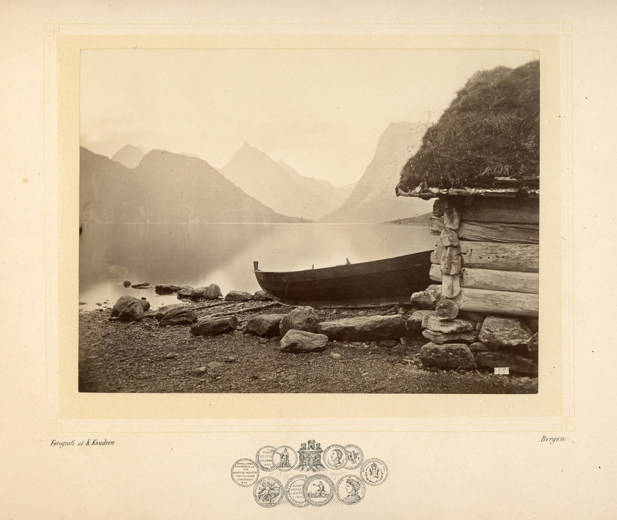 Motiv av en robåt i støa ved et lafta naust. I bakgrunnen er ...