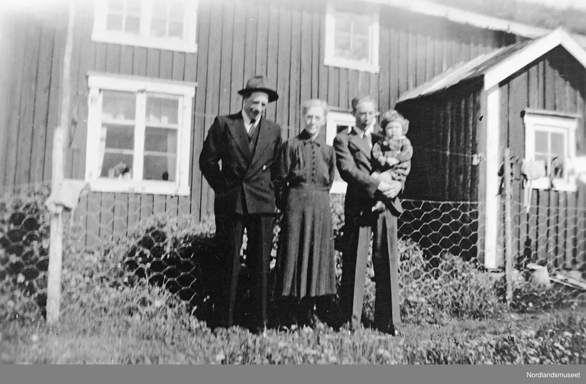 Tre voksne, en av dem holder et barn. De står foran et hus med ...