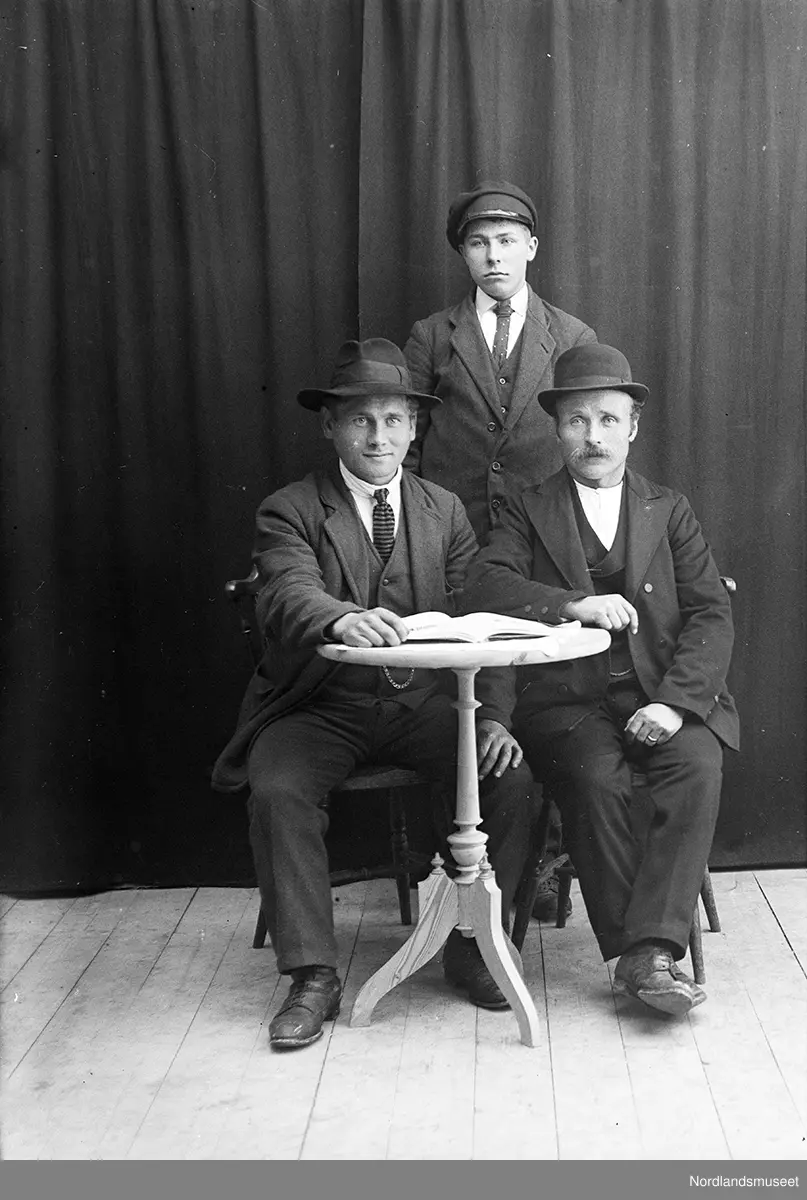 Portrett av tre menn. To sitter på hver sin stol bak et lite rundt bord ...